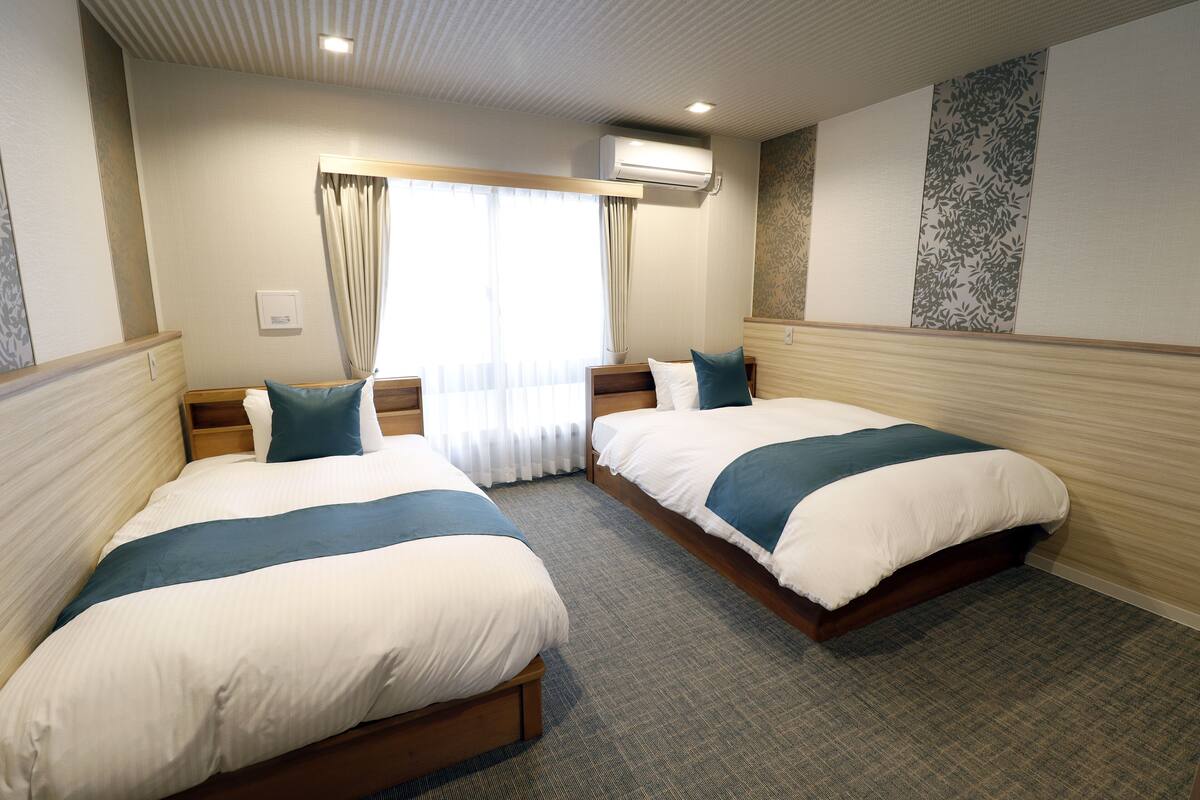 Quarto de hotel com duas camas, uma janela com cortinas, um ar condicionado e uma parede estampada.