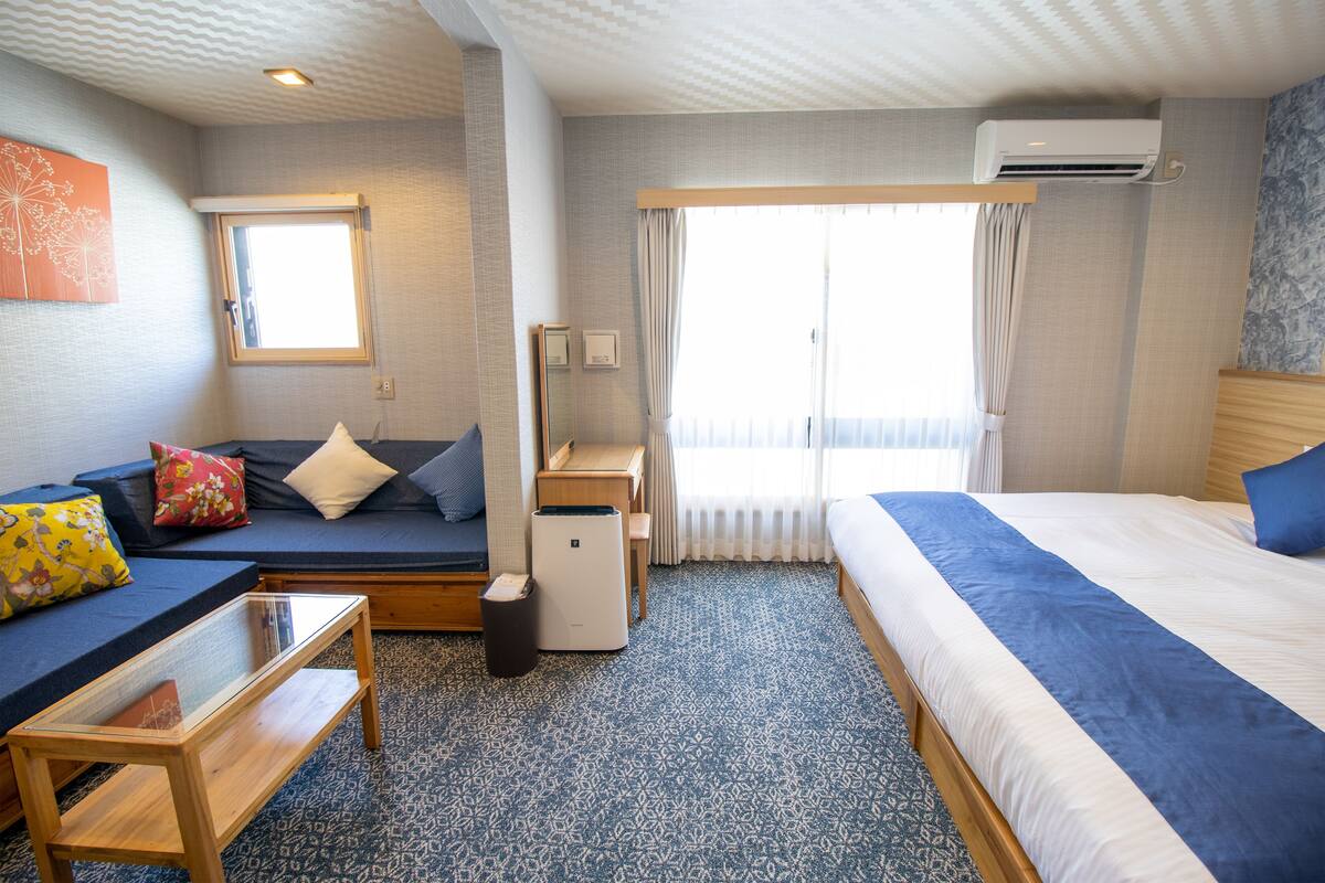 Quarto de hotel com cama, sofá, mesa pequena, escrivaninha, ar condicionado e janela com cortinas.