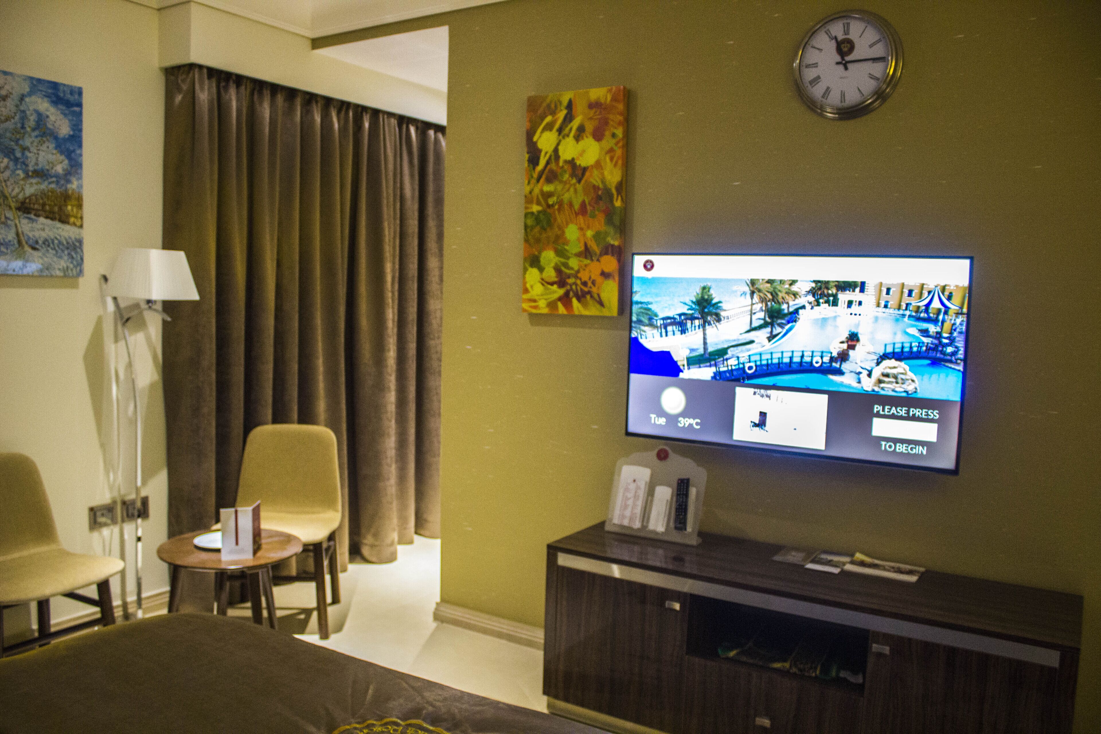 Quarto de hotel com cama, televisão na parede, relógio, pintura e janela com cortinas.