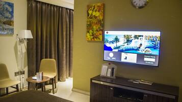 Quarto de hotel com cama, televisão na parede, relógio, pintura e janela com cortinas.