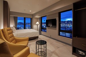 Quarto de hotel moderno com uma cama grande, um espaço de estar com uma poltrona, uma mesa pequena e uma televisão.