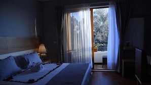 Quarto de hotel com cama, mesa de cabeceira, luminária e vista para o exterior através de uma janela com cortinas.