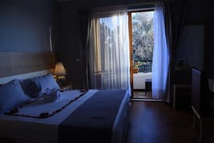 Quarto de hotel com cama, mesa de cabeceira, luminária e vista para o exterior através de uma janela com cortinas.