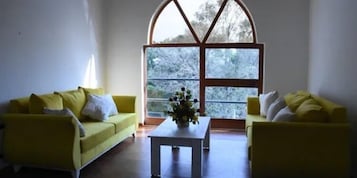 Quarto com dois sofás amarelos, uma mesa de centro branca e uma janela grande com vista para árvores.
