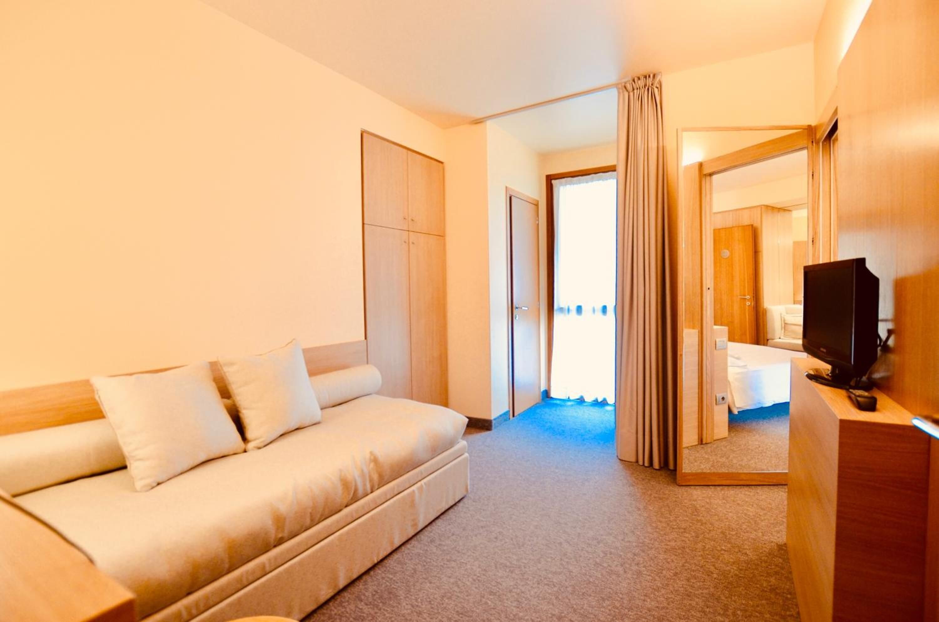 Quarto de hotel com sofá, televisão sobre suporte de madeira e porta para uma varanda.