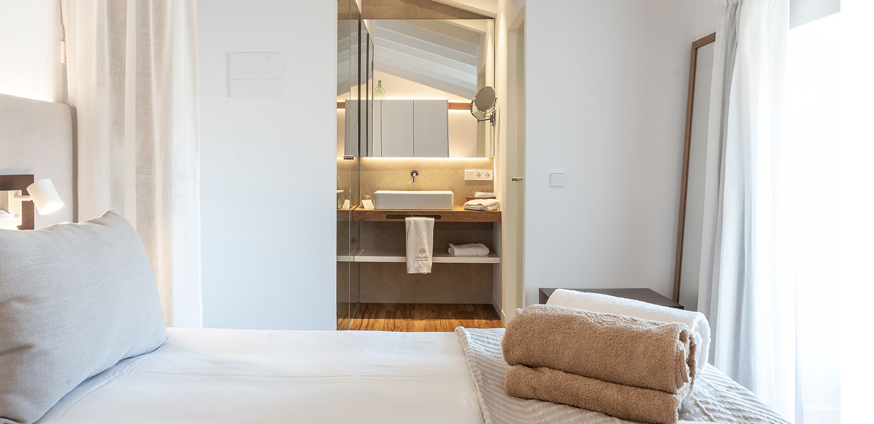 Quarto de hotel com cama, mesa de cabeceira, espelho, banheiro com pia e porta-toalhas.