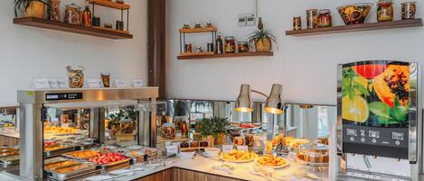 Uma área de buffet com diversos alimentos, uma máquina de café e prateleiras com potes e plantas.