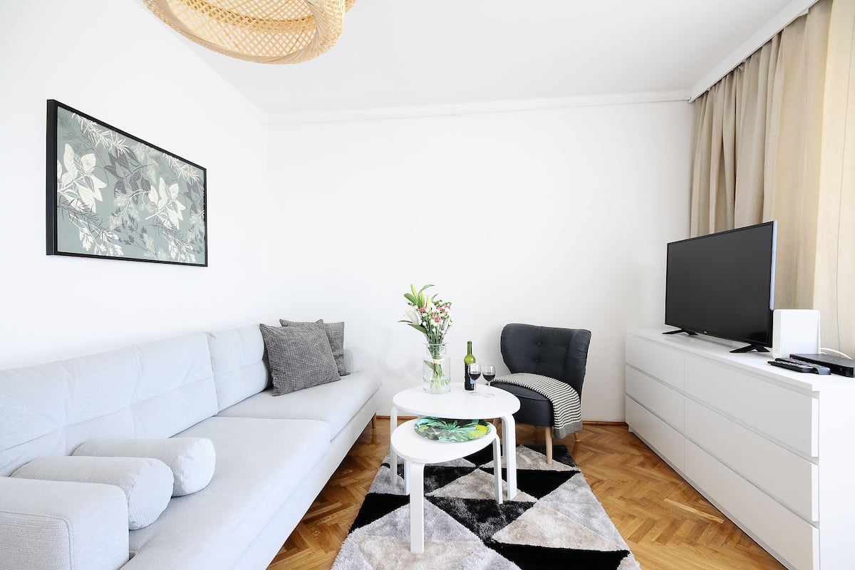 Uma sala de estar moderna com um sofá cinza, uma mesa de centro branca com um vaso de flores, uma televisão de tela plana em um armário branco e um tapete preto e branco.