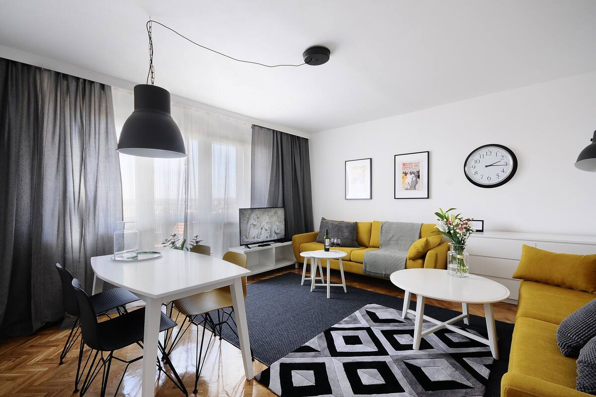 Uma sala de estar moderna com uma mesa branca, cadeiras pretas, um sofá amarelo e uma luminária pendente preta.