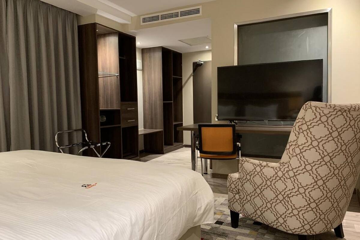 Quarto de hotel com uma cama grande, televisão, guarda-roupa e um espaço de estar com uma poltrona estampada.