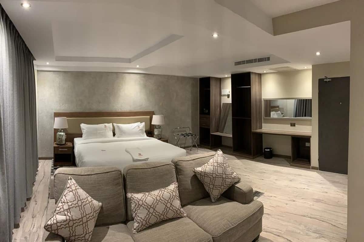 Quarto de hotel moderno com uma cama grande, um sofá com almofadas estampadas, uma mesa de cabeceira com abajur, um guarda-roupa e um espelho.
