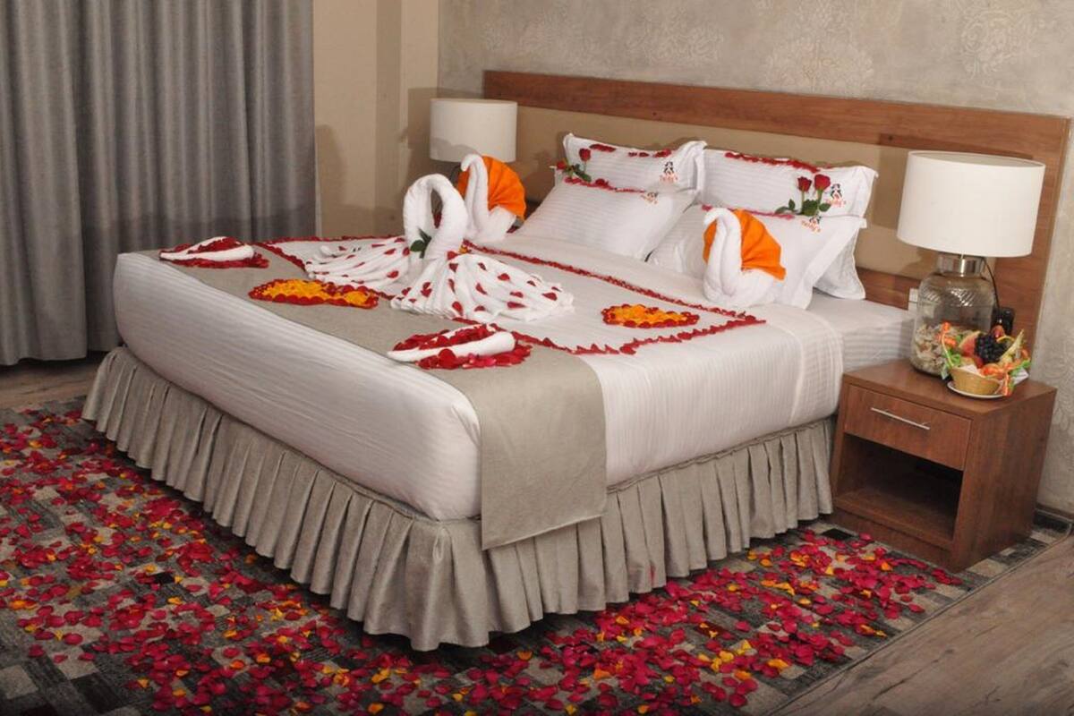 Uma cama bem arrumada com pétalas de rosa e decorações de cisne, um criado-mudo de madeira com abajur e um tapete estampado.
