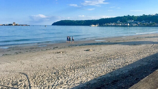 Uma praia com areal dourado e mar calmo. Algumas pessoas caminham pela areia. Ao fundo, vislumbra-se uma cidade com edifícios e uma colina coberta de árvores.