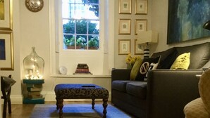 Sala de estar com assoalho de madeira, um banquinho estampado, um sofá de couro escuro com almofadas amarelas, uma janela com vista para vegetação e quadros nas paredes.