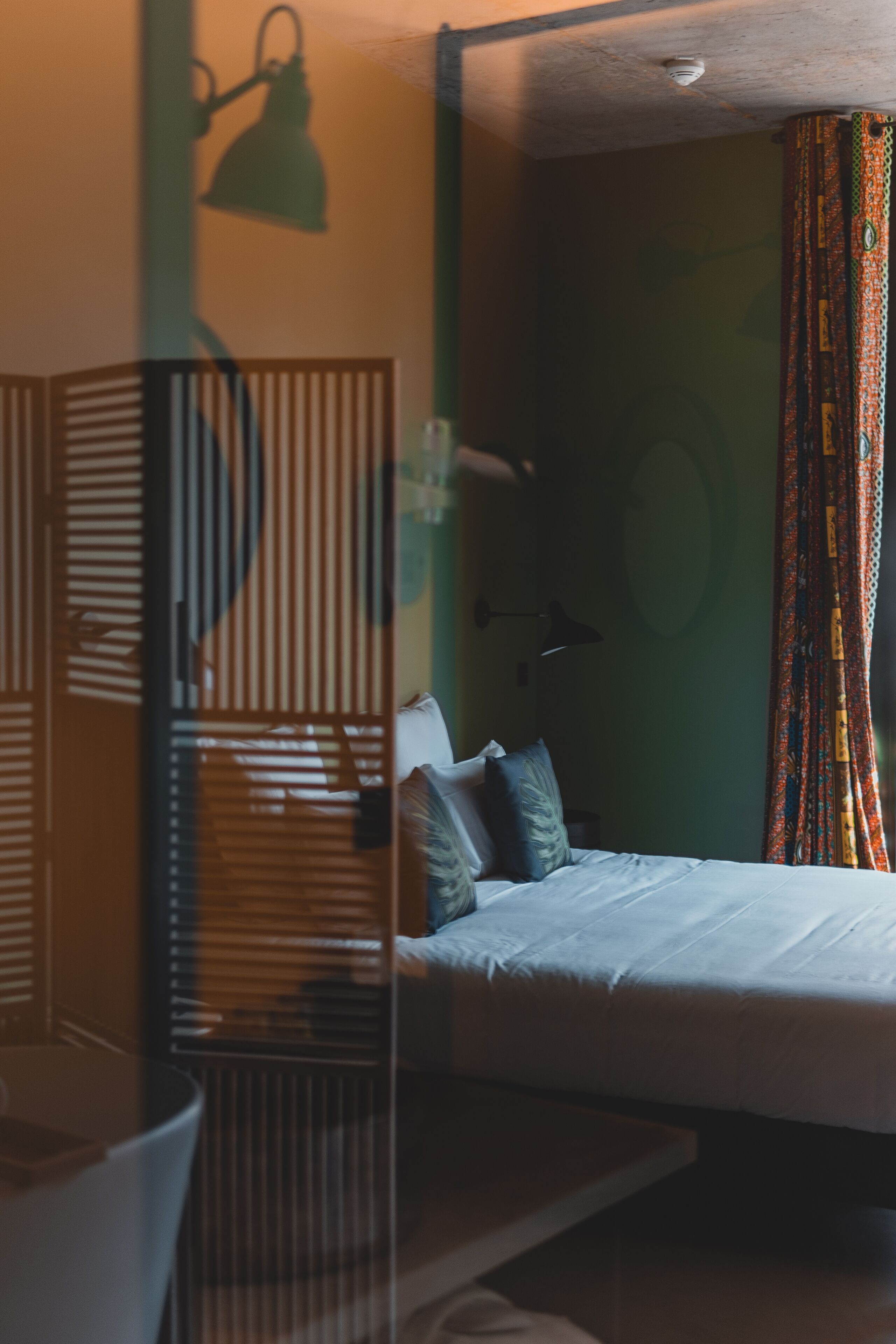 Quarto com cama, mesa de cabeceira, abajur e janela com cortinas.