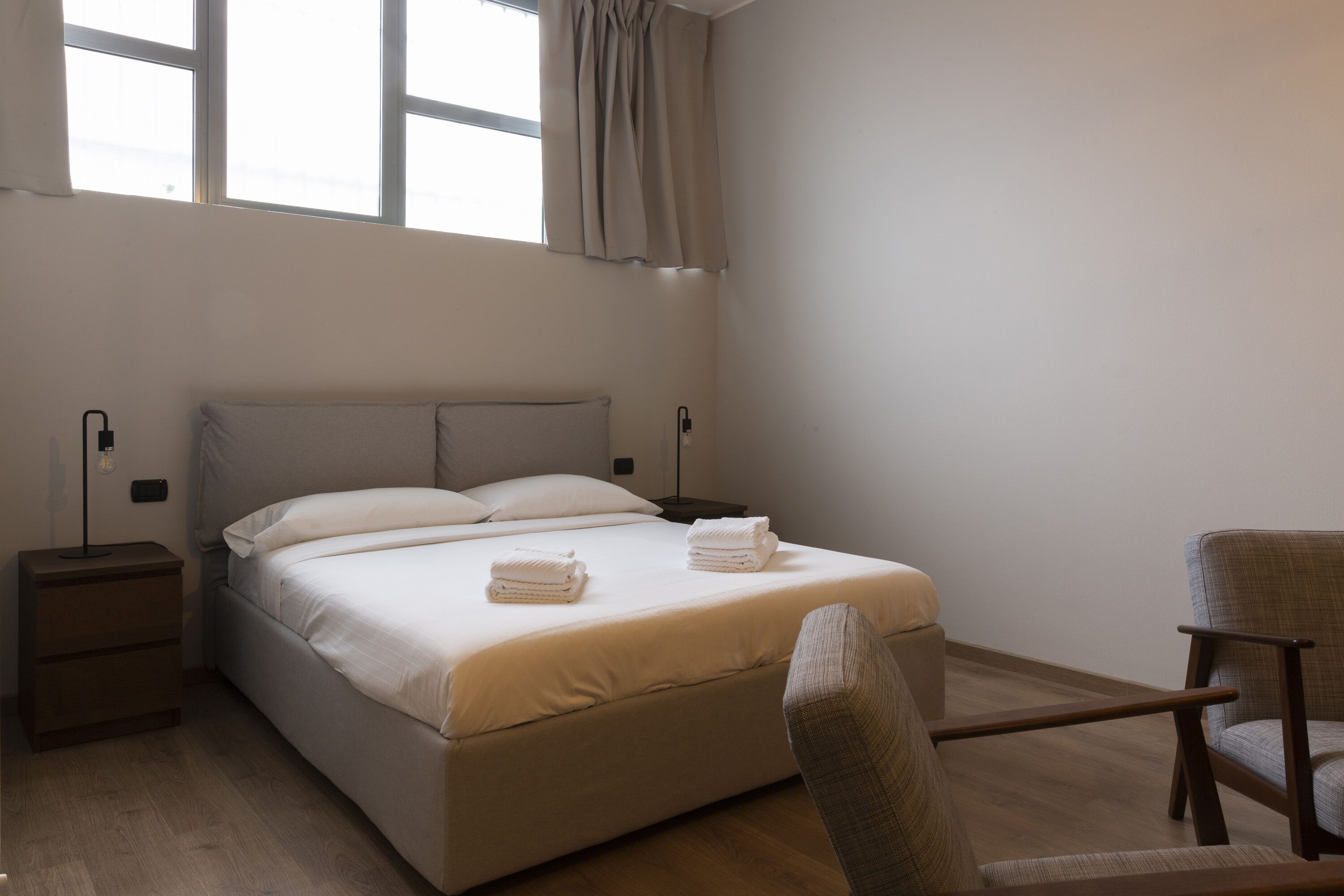 Quarto com cama, duas mesas de cabeceira, uma cadeira e uma janela com cortinas.