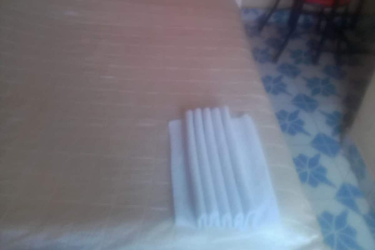 Uma cama com cabeceira vermelha, colchão branco e uma toalha branca dobrada. Há uma cadeira com almofada vermelha e uma mesa com toalha de mesa estampada em azul.