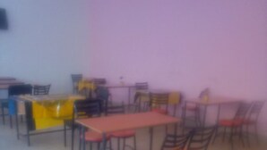 Uma área de refeições com mesas e cadeiras, uma toalha de mesa amarela em uma das mesas e uma parede rosa.