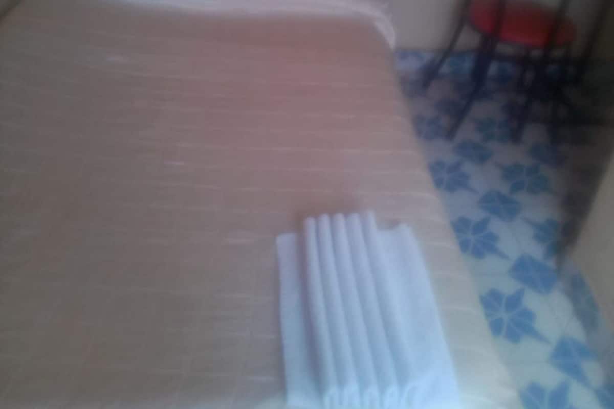 Cama com cabeceira vermelha, colchão branco e edredom azul com padrão floral. Há uma cadeira com almofada vermelha e uma mesa com toalha de mesa azul estampada com flores.