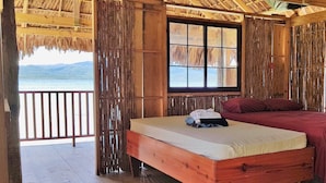 Uma cama de madeira com colchão e almofadas, uma janela com vista para o mar e uma cortina de bambu.