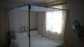 Uma cama de dossel com cortina, um ventilador na mesa de cabeceira e uma janela com cortinas leves.