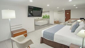 Quarto de hotel moderno com cama, televisão, sofá, poltrona, secretária, planta e banheira.