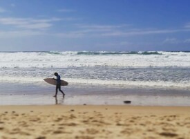 Cena de praia com ondas e uma pessoa carregando uma prancha de surf.