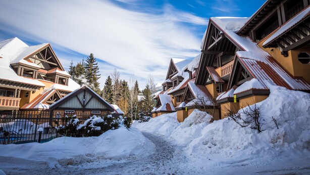 Uma trilha coberta de neve que leva a uma fileira de edifícios tradicionais em estilo chalé, com telhados inclinados e fachadas de madeira.