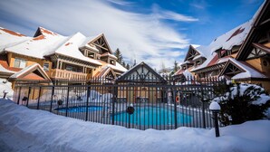 Piscina ao ar livre coberta de neve, rodeada por edifícios tipo chalé, com um céu azul límpido.