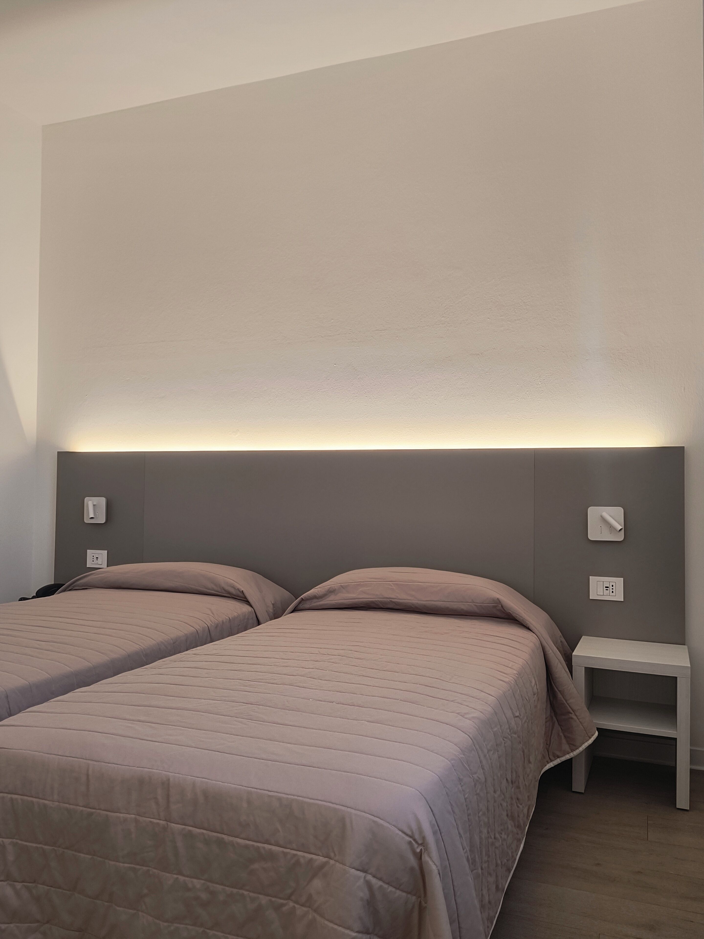 Quarto de hotel moderno com duas camas, uma mesa de cabeceira e luminária fixada na parede.