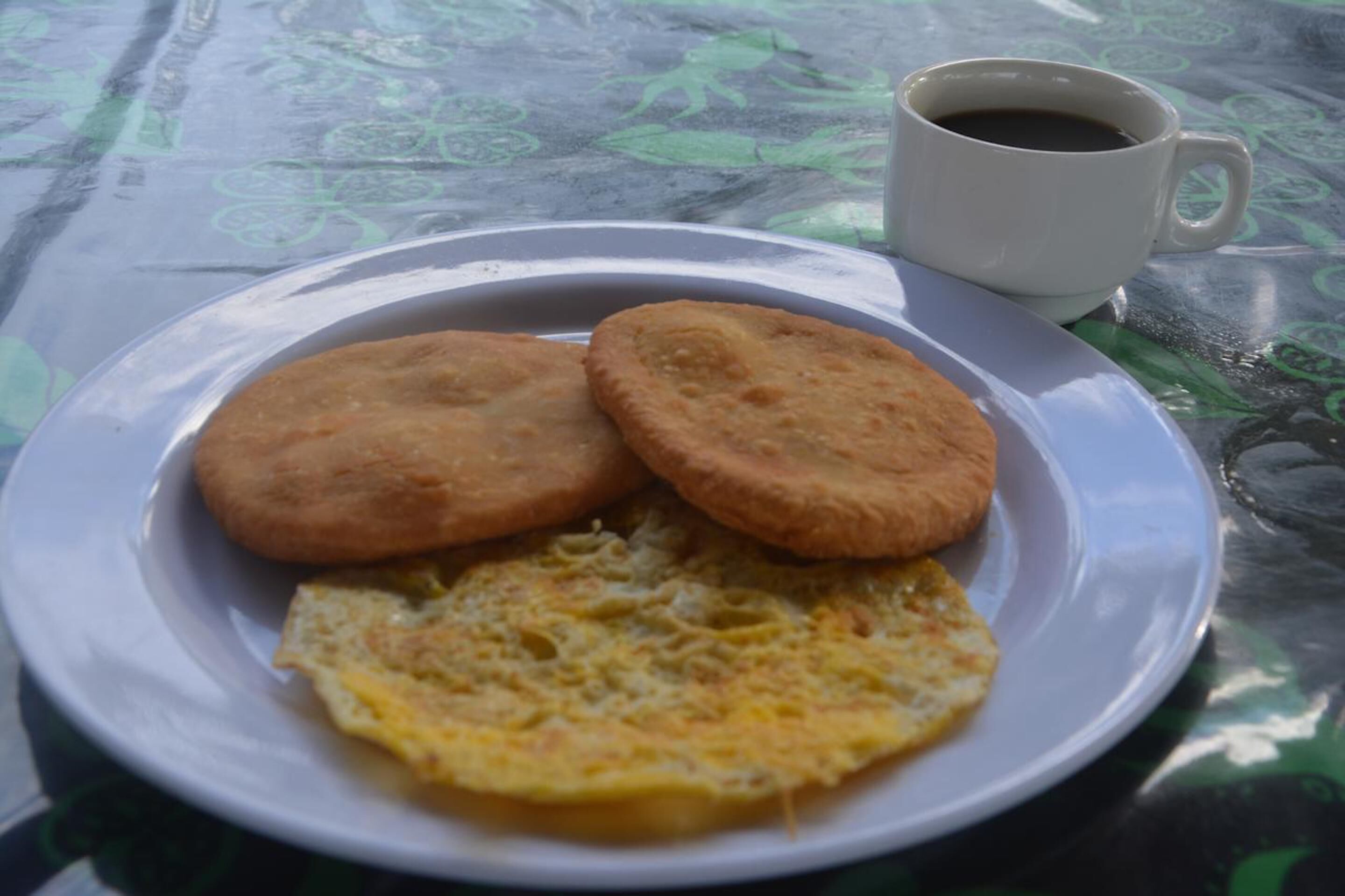 Um prato com dois alimentos fritos e um omelete, acompanhado por uma xícara de café sobre uma mesa com toalha de mesa estampada.