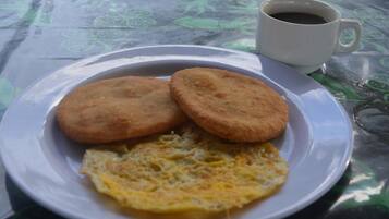Um prato com dois alimentos fritos e um omelete, acompanhado por uma xícara de café sobre uma mesa com toalha de mesa estampada.