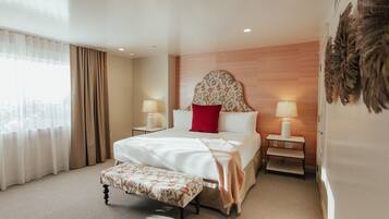 Quarto de hotel com uma cama grande, uma mesa de cabeceira com luminária, um banco e vista para a cidade pela janela.