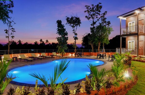 Piscina com espreguiçadeiras e mesas, rodeada por árvores e um edifício com varanda.