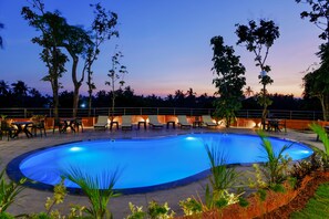 Uma piscina com espreguiçadeiras e mesas, cercada por árvores e plantas.