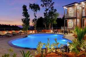 Piscina com espreguiçadeiras e mesas, cercada por plantas e um edifício com varandas.