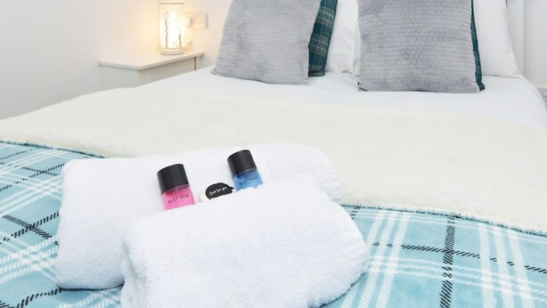 Cama bem arrumada com manta branca e colcha xadrez. Há duas embalagens de loção sobre a cama.