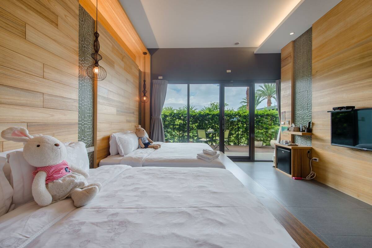 Quarto de hotel com duas camas, televisão, pequena cozinha e varanda com vista para vegetação.