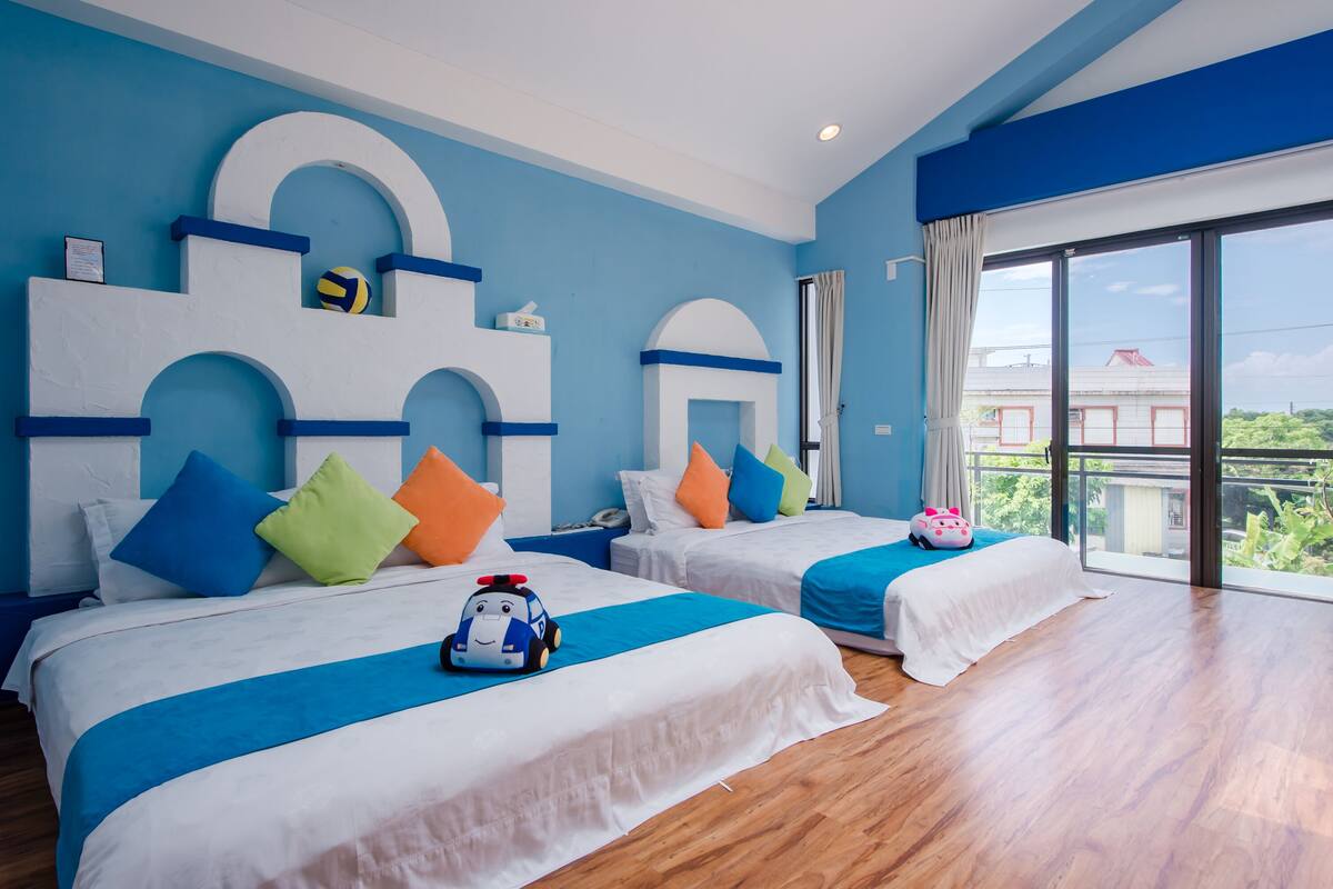 Quarto de hotel com duas camas, almofadas coloridas, um carro de polícia de brinquedo e vista para o verde através de grandes janelas.