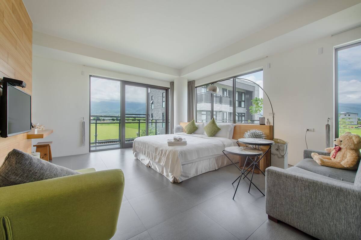 Quarto de hotel moderno com uma cama grande, um sofá, uma mesinha e vista da cidade por grandes janelas.
