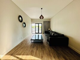 Uma sala de estar ampla com piso de madeira, um sofá preto, uma mesa de centro de vidro e uma luminária pendente.