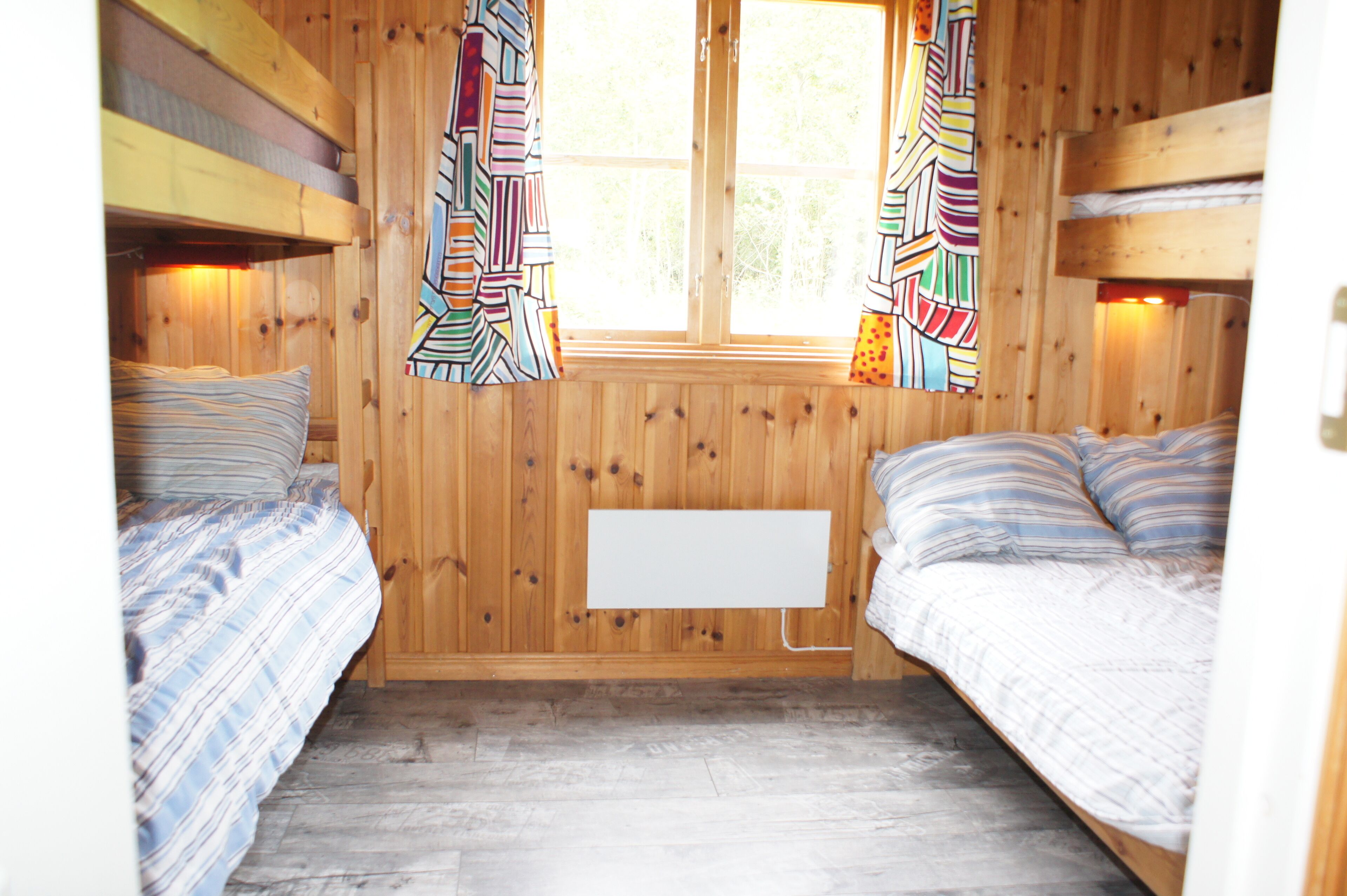 Um quarto com duas camas beliche, paredes de madeira, uma janela com cortinas e um radiador.