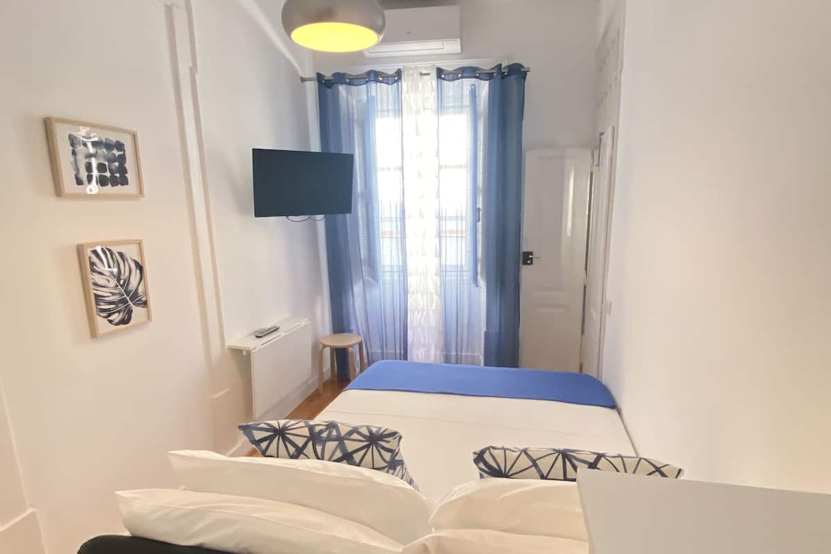 Um quarto com uma cama, uma escrivaninha, uma cadeira, uma televisão e uma janela com cortinas.