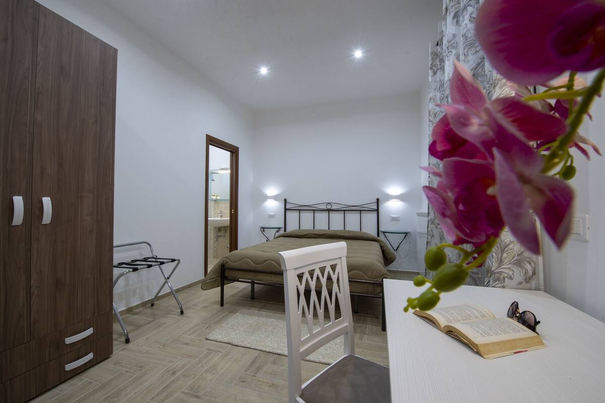 Quarto com cama, guarda-roupa, cadeira, mesa com um livro e óculos, e um espelho.