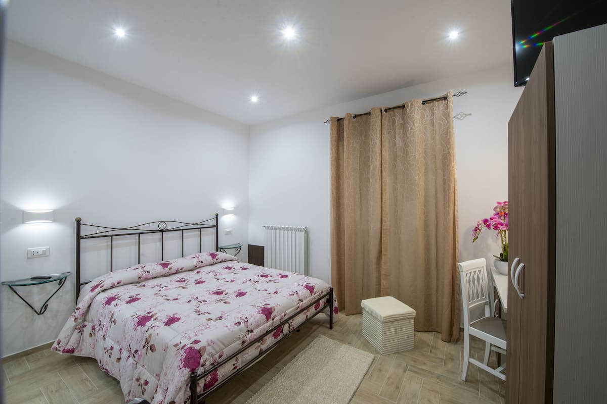 Quarto com cama, mesa de cabeceira, abajur e janela com cortinas.