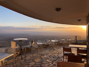 Terraço ao ar livre com mesas e cadeiras, com vista para uma paisagem montanhosa ao pôr do sol.
