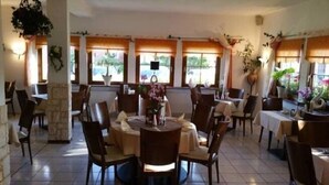 Uma sala de jantar com várias mesas e cadeiras, janelas amplas com cortinas e plantas em vasos.