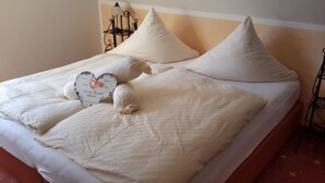 Uma cama bem arrumada com lençóis brancos, um travesseiro decorativo em formato de coração e uma luminária de mesa.