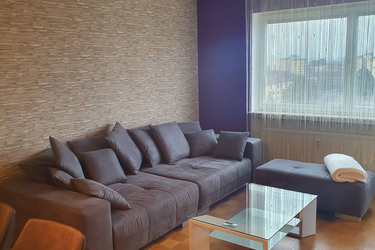 Uma sala de estar moderna com uma parede texturizada em pedra, um sofá cinza com várias almofadas, uma mesa de centro de vidro e um ventilador de teto.