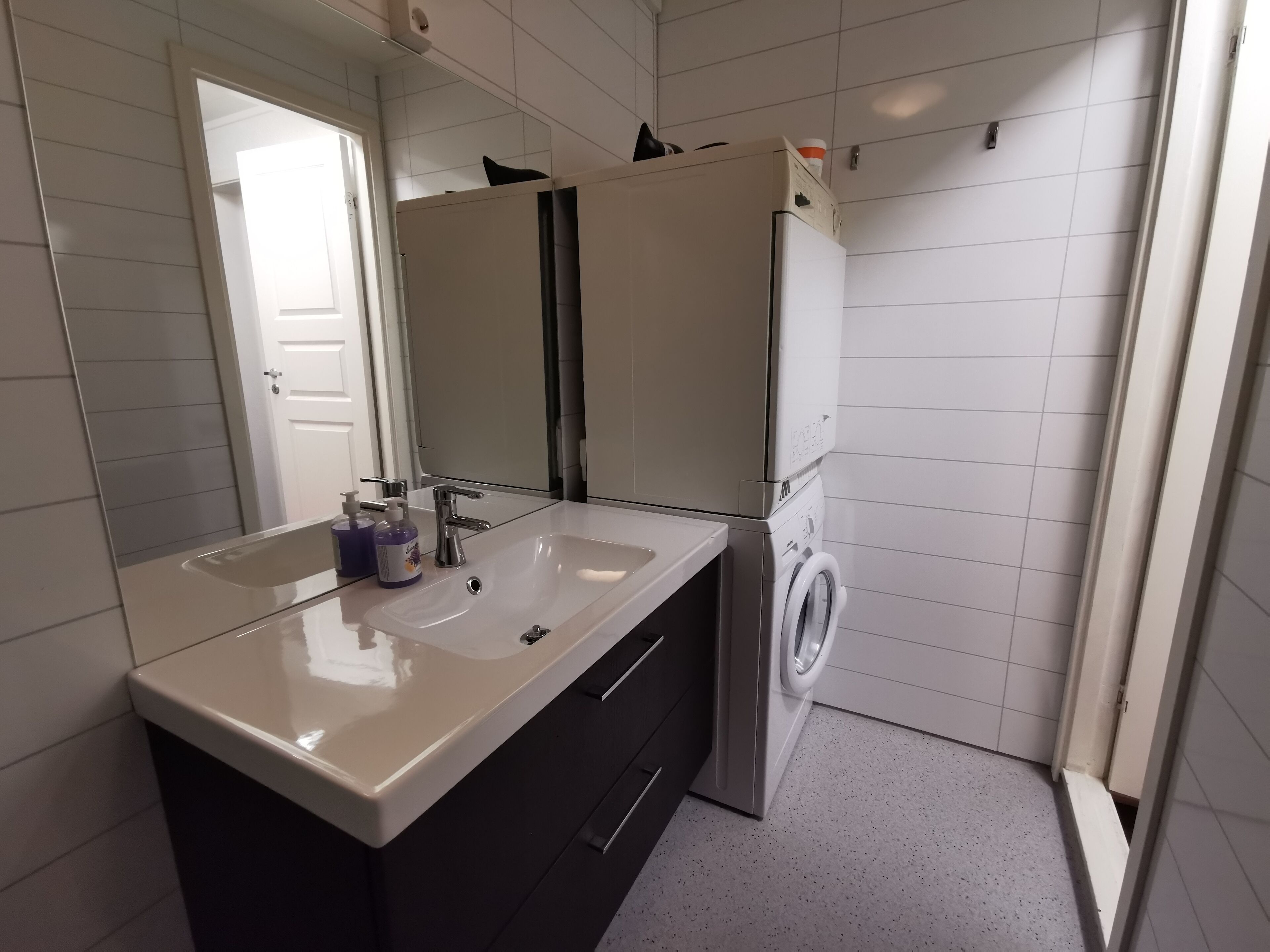 Casa de banho moderna com lavatório duplo, espelho, máquina de lavar roupa e porta para outro espaço.